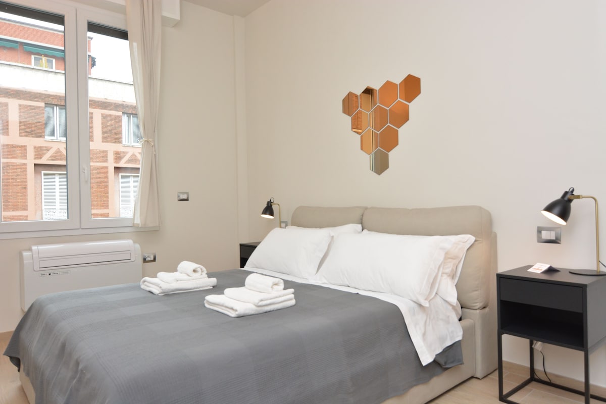 Quarto de hotel com cama, abajures, aquecedor, janela e um objeto decorativo na parede.