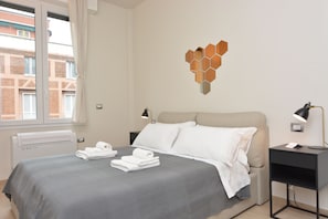 Quarto de hotel com cama, abajures, aquecedor, janela e um objeto decorativo na parede.