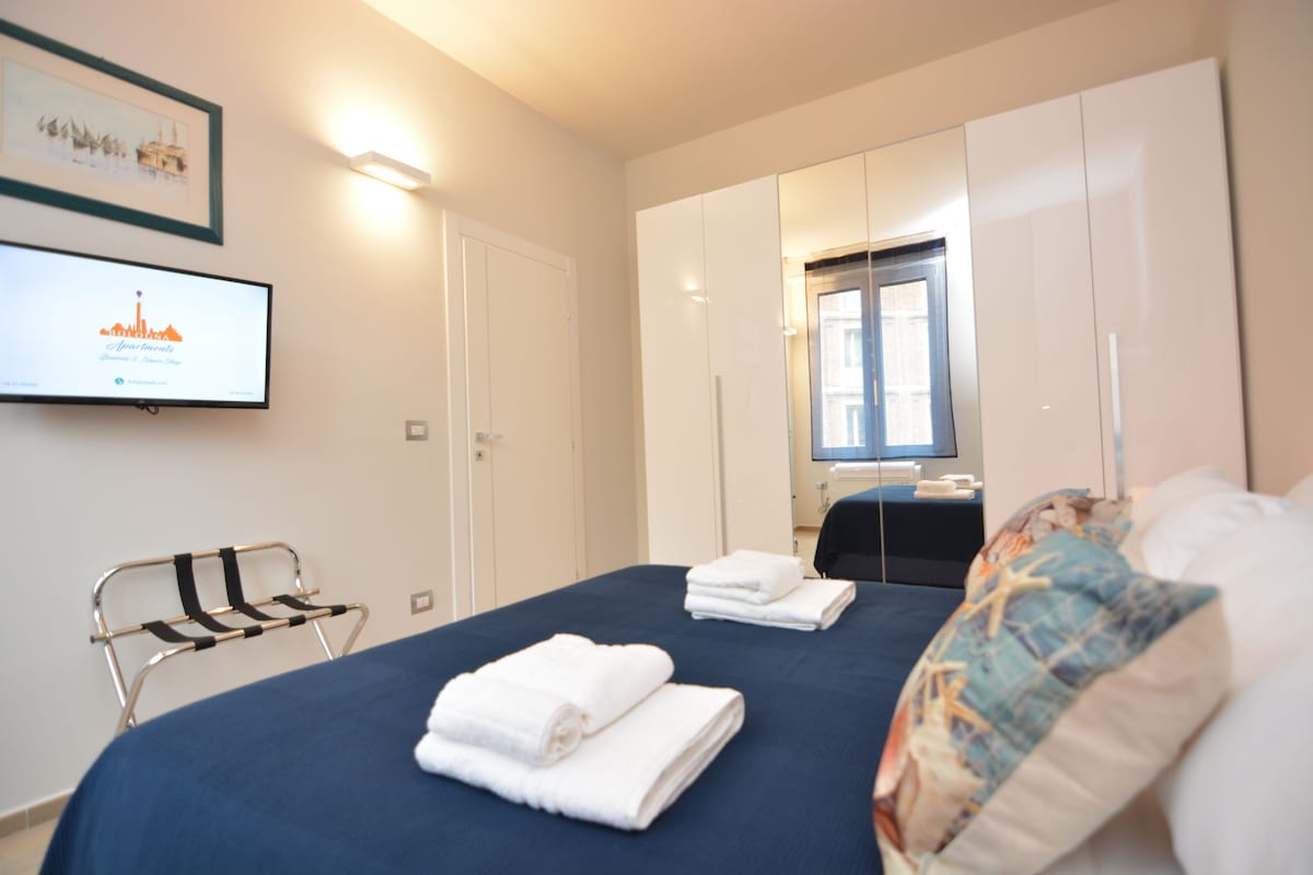 Quarto de hotel com cama, cadeira, escrivaninha, janela e televisão fixada na parede exibindo uma vista urbana.