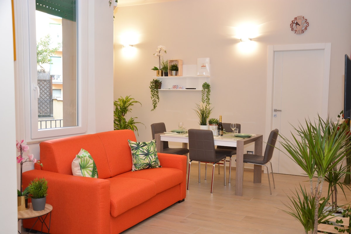 Uma sala de estar moderna com um sofá laranja vibrante, uma mesa de jantar com cadeiras e diversos vasos de plantas.