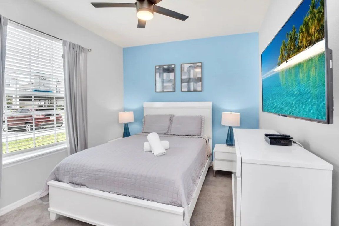 Quarto com cama, mesas de cabeceira, ventilador de teto, janela com persianas e um quadro de uma praia.