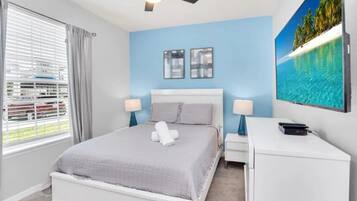 Quarto com cama, mesas de cabeceira, ventilador de teto, janela com persianas e um quadro de uma praia.