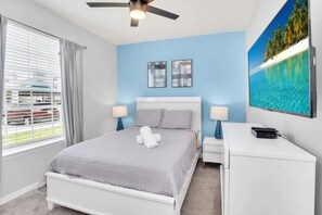 Quarto com cama, mesas de cabeceira, ventilador de teto, janela com persianas e um quadro de uma praia.