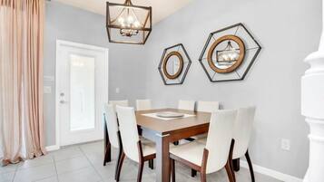 Uma sala de jantar com mesa de madeira e cadeiras brancas, um lustre e dois espelhos hexagonais na parede.