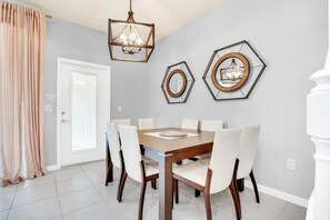 Uma sala de jantar com mesa de madeira e cadeiras brancas, um lustre e dois espelhos hexagonais na parede.