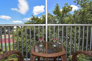 Varanda com mesa de vidro e cadeiras de vime, com vista para uma quadra de tênis.