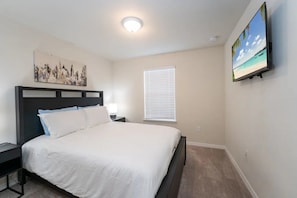 Quarto com cama, mesa de cabeceira, TV fixada na parede e janela com persianas.