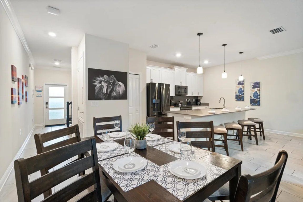 Área de refeições com mesa posta para quatro pessoas, cozinha ao fundo e iluminação pendente.