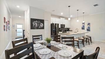 Área de refeições com mesa posta para quatro pessoas, cozinha ao fundo e iluminação pendente.