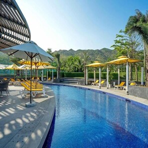 Piscina com espreguiçadeiras e guarda-sóis, rodeada por vegetação e colinas.