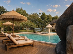 Área de piscina com espreguiçadeiras, um guarda-sol de palha e uma escultura de pedra.