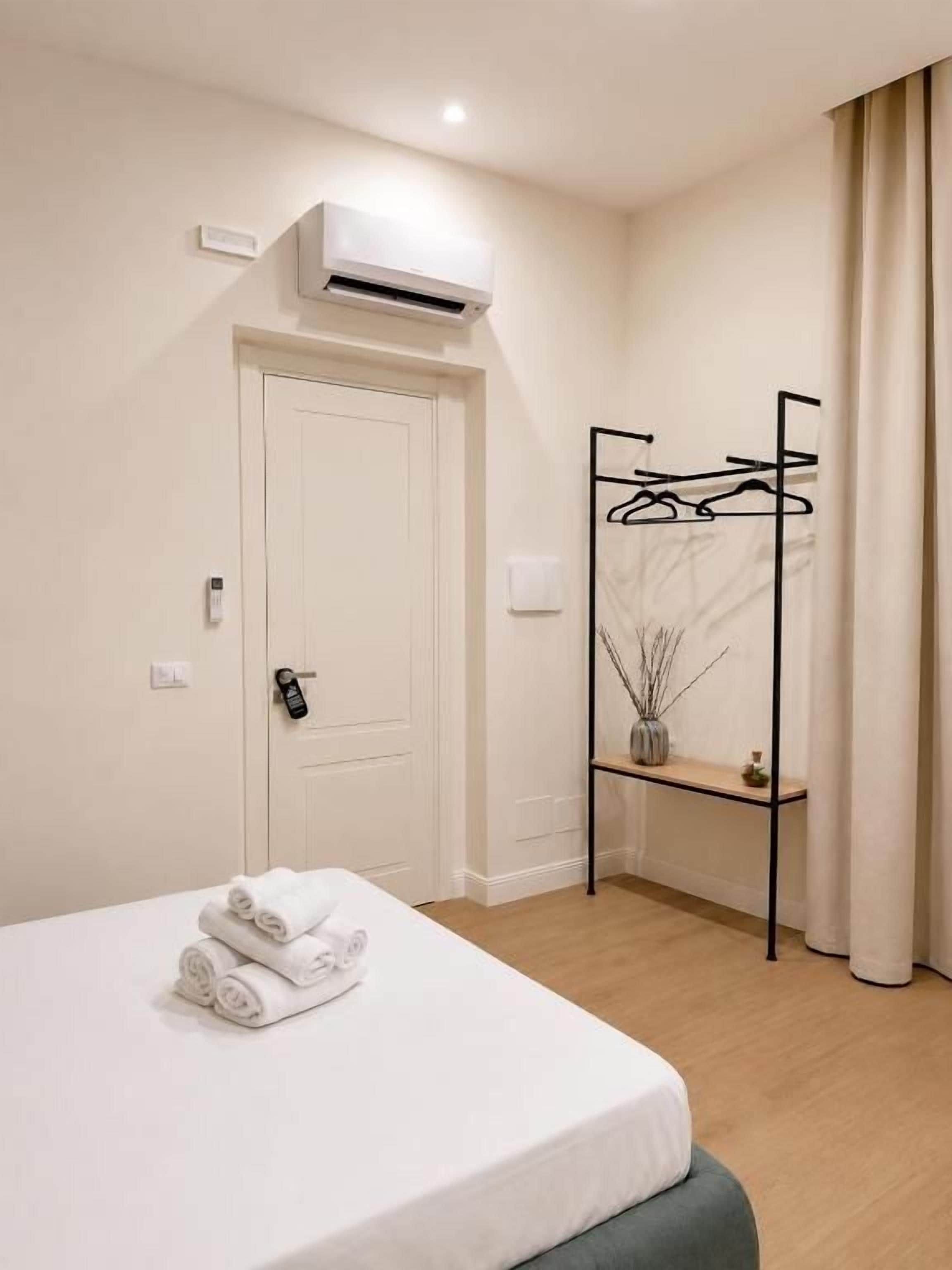 Quarto de hotel com cama, porta branca, unidade de ar condicionado, cabideiro fixado na parede e pequena mesa com um vaso.