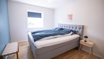 Quarto com cama, mesa de cabeceira, abajur, janela e um pequeno banco.