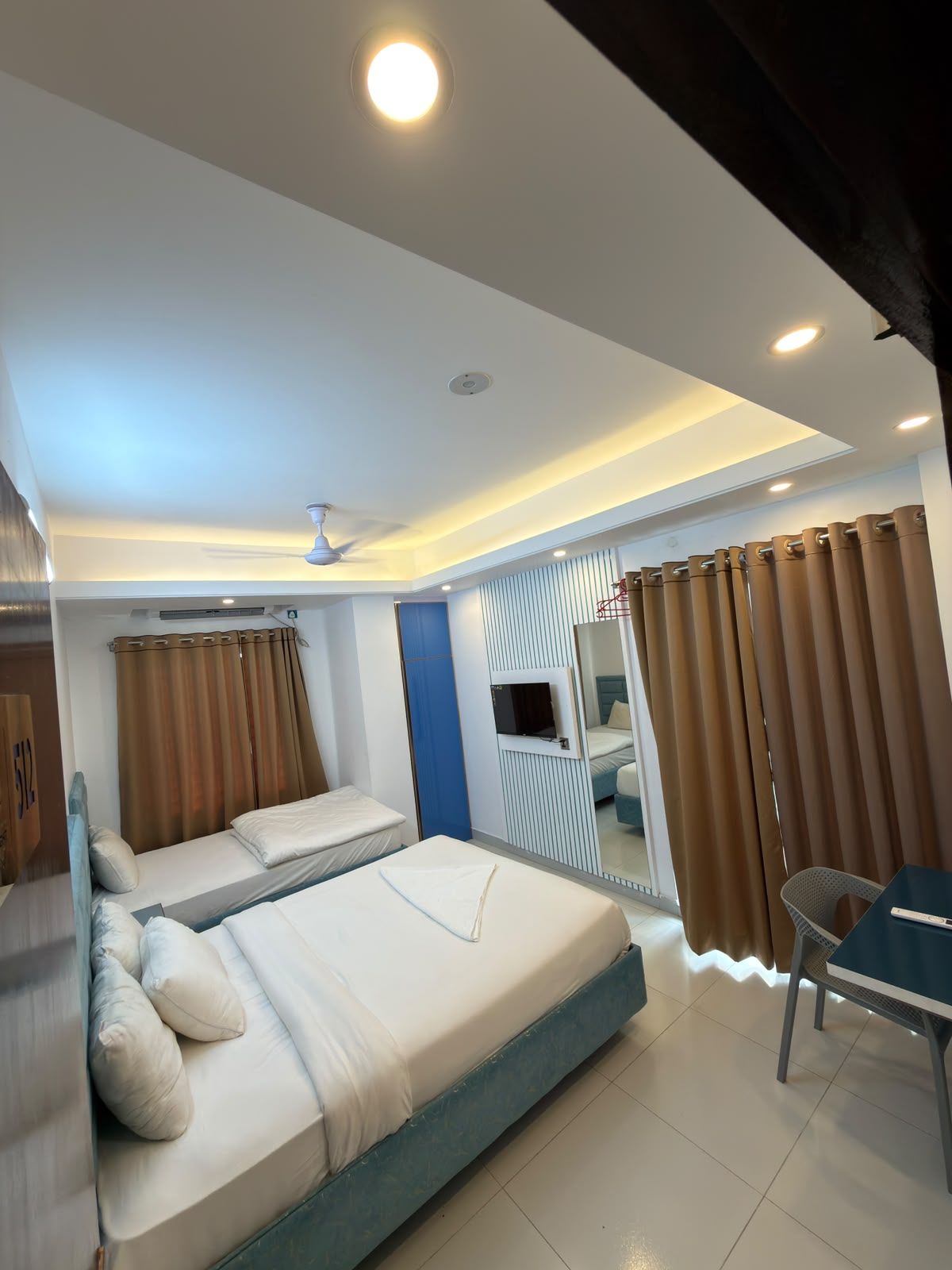 Quarto de hotel com duas camas, uma escrivaninha, uma cadeira, ventoinha no teto e televisão.