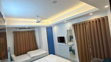 Quarto de hotel com duas camas, uma escrivaninha, uma cadeira, ventoinha no teto e televisão.
