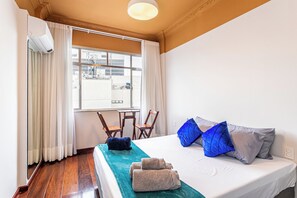 Quarto de hotel com uma cama, duas cadeiras, uma janela com cortinas e uma luminária no teto.
