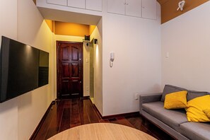 Um corredor com porta de madeira, TV de tela plana fixada na parede, sofá com almofadas amarelas e piso de madeira.