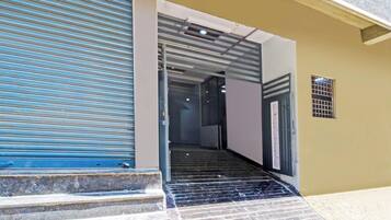 Entrada de um edifício com uma persiana azul fechada, escadas que levam a um andar superior e uma pequena janela com padrão de grade.