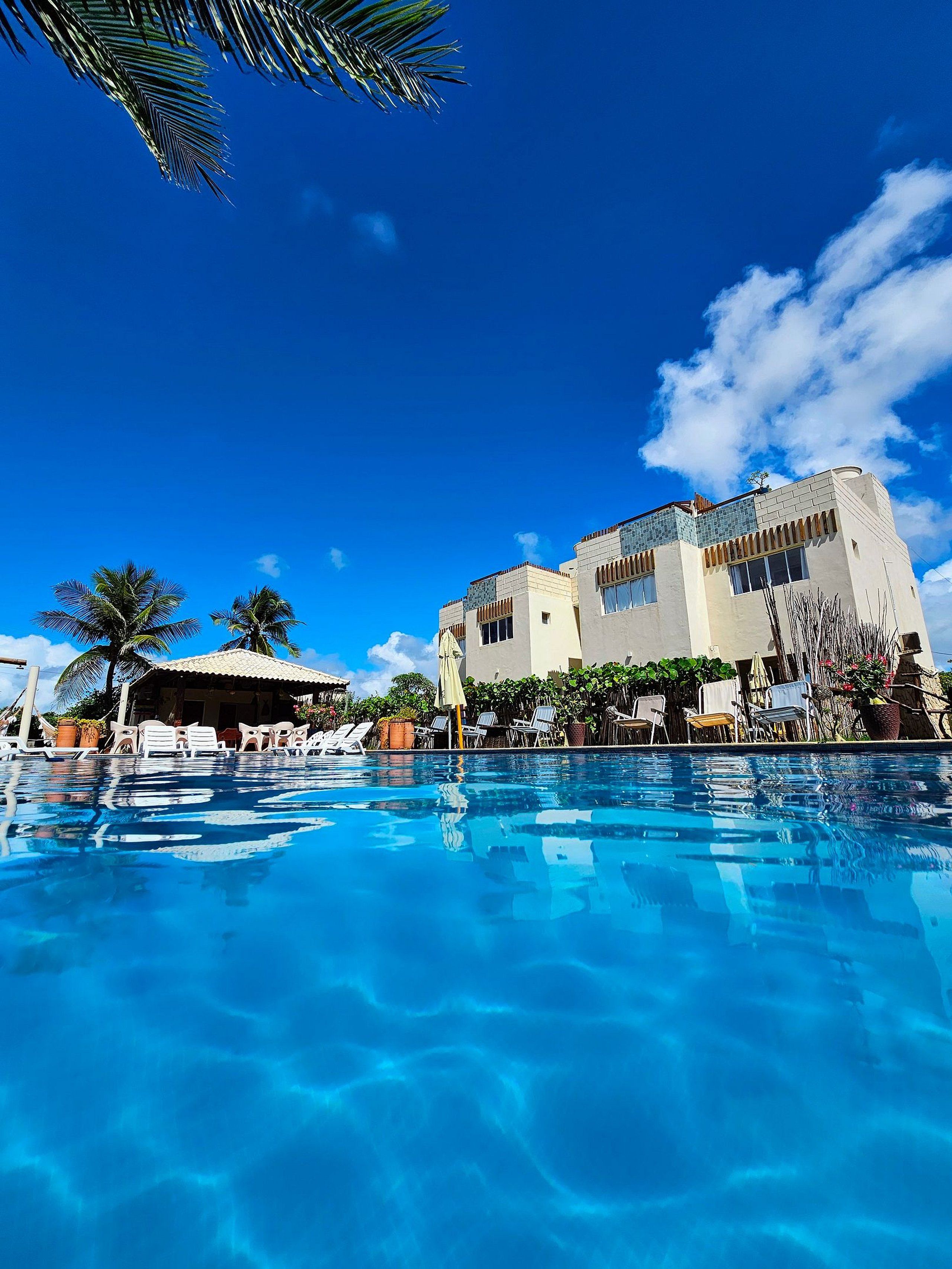 Piscina azul límpida com espreguiçadeiras e guarda-sóis, um edifício com telhado de azulejos e palmeiras.