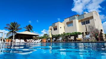 Piscina azul límpida com espreguiçadeiras e guarda-sóis, um edifício com telhado de azulejos e palmeiras.