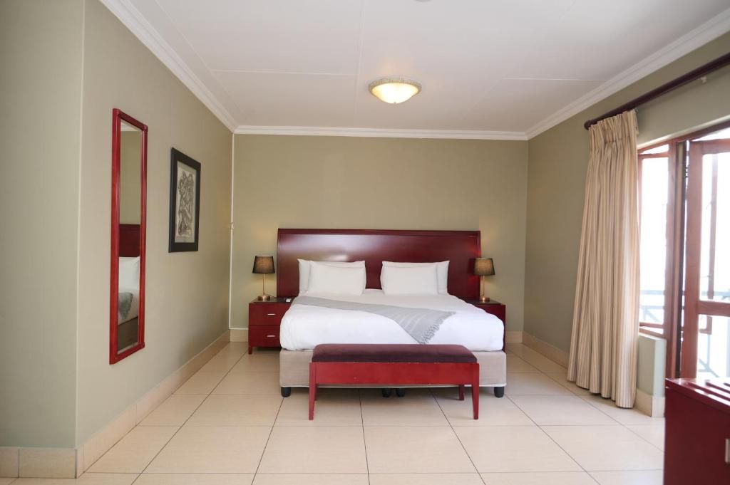 Quarto de hotel com uma cama grande, mesas de cabeceira, um banco, um espelho e uma janela com cortinas.