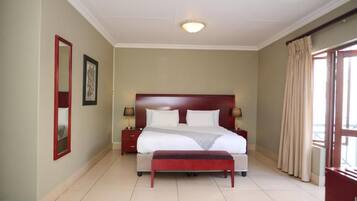 Quarto de hotel com uma cama grande, mesas de cabeceira, um banco, um espelho e uma janela com cortinas.