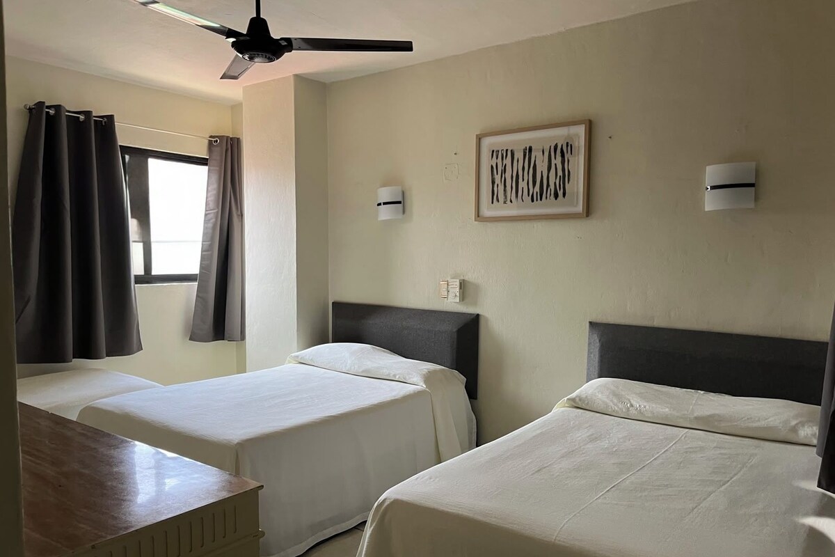 Quarto de hotel com duas camas de solteiro, um ventilador de teto, janela com cortinas e uma moldura com quadro na parede.