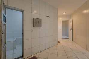 Um corredor com paredes e chão de azulejos brancos, uma porta à esquerda e um interruptor de luz na parede.