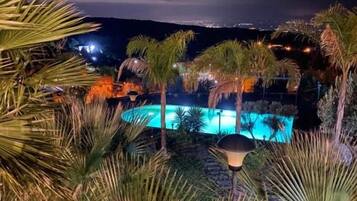 Vista noturna de uma área de piscina com palmeiras e piscina iluminada.