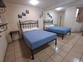 Quarto com duas camas, uma mesinha e um ventilador fixado na parede.
