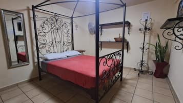 Uma cama de dossel com colcha vermelha, um espelho na parede, um ventilador e um vaso de plantas.
