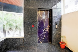 Porta de elevador de mármore com superfície refletora, um botão de emergência vermelho e um vaso de planta ao lado.