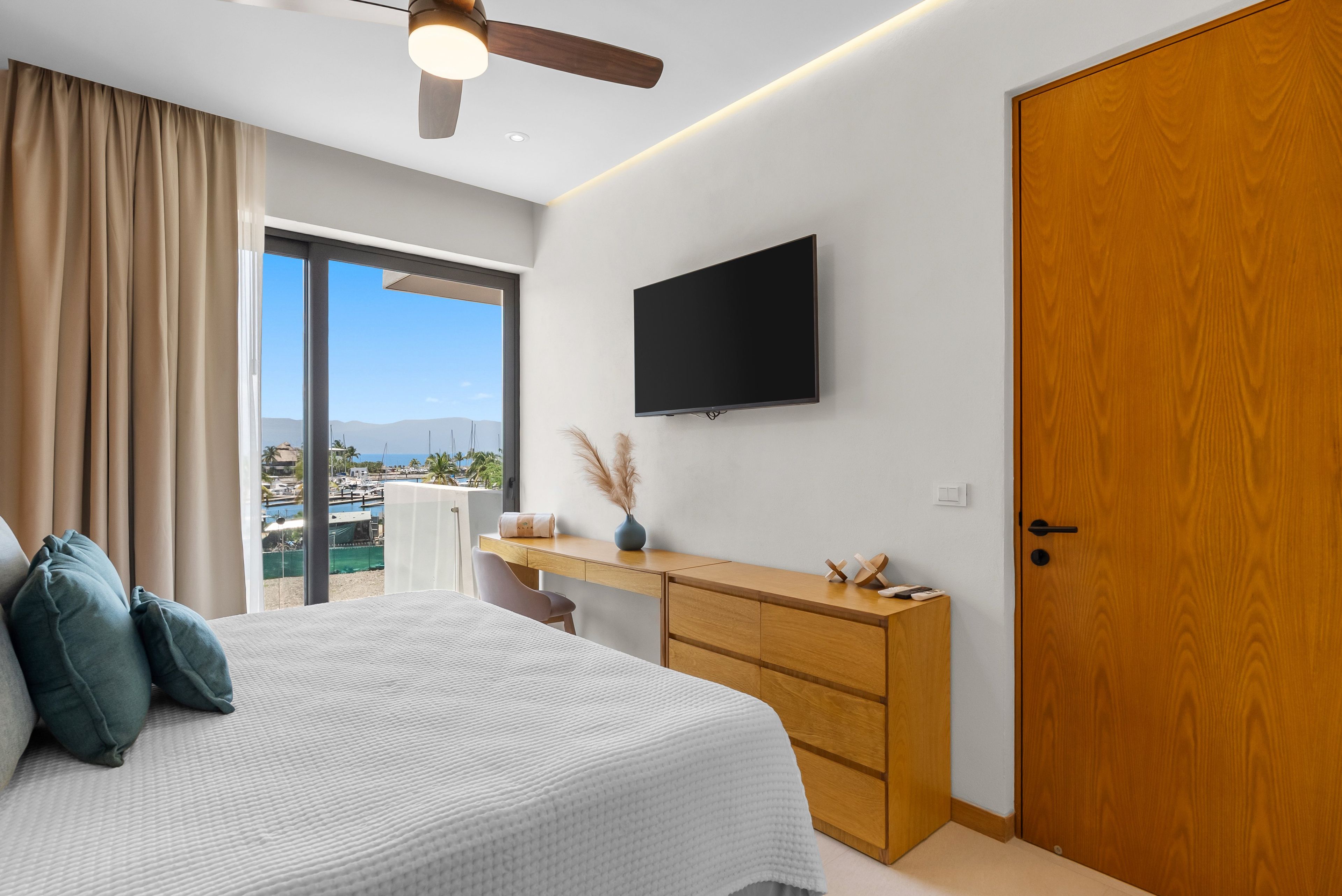 Quarto com cama, escrivaninha, cadeira, televisão e vista para o exterior.