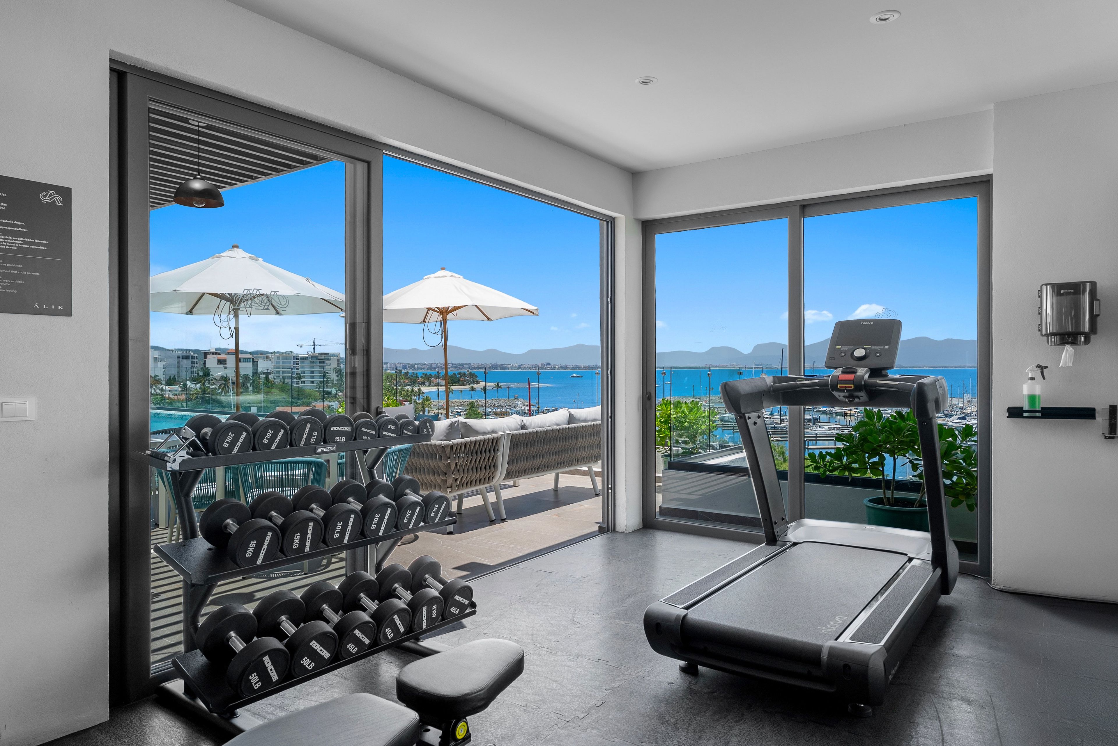 Academia moderna com esteira, halteres e equipamentos de ginástica, com vista panorâmica para o mar e a paisagem urbana.