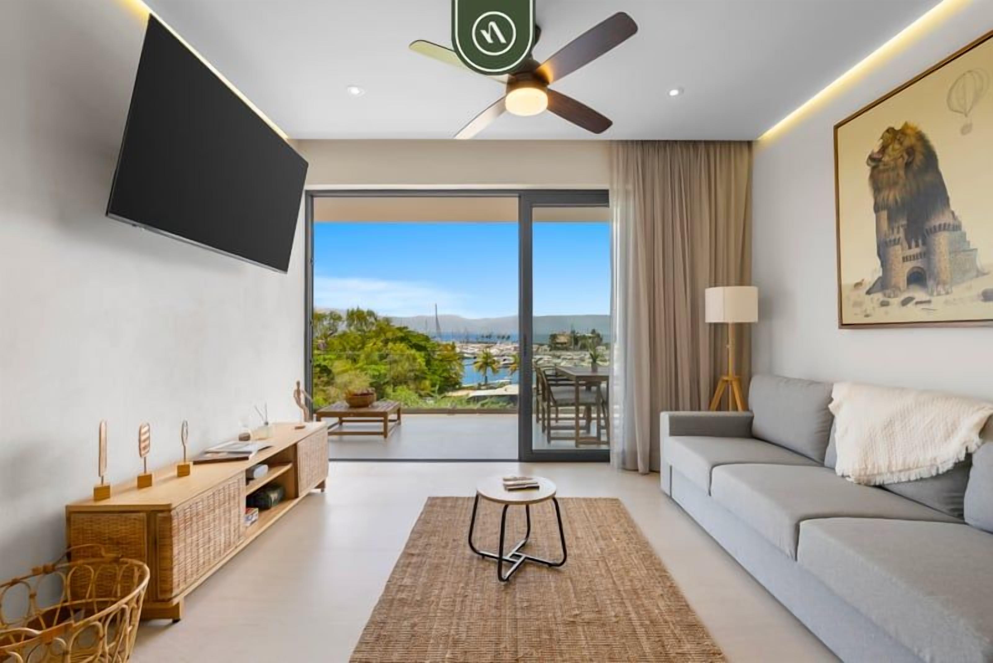 Uma sala de estar moderna com televisão de tela plana, ventilador de teto, sofá, mesa de centro e vista para o exterior.