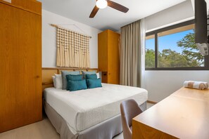 Quarto com cama, almofadas, cabeceira de madeira, ventoinha no teto, janela com cortinas e mesa de madeira.