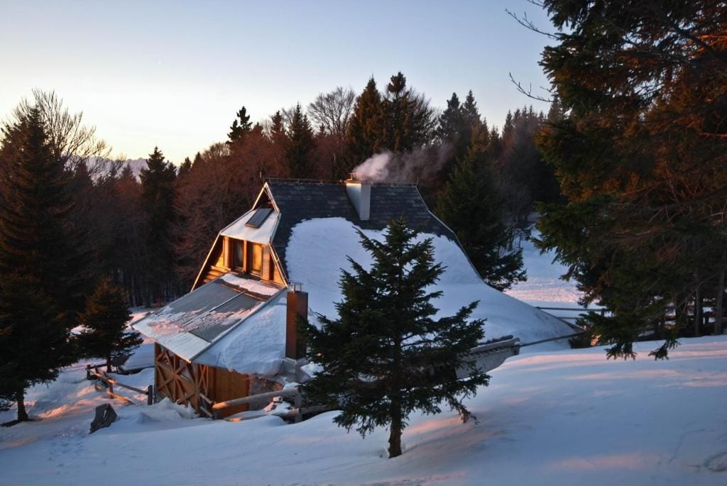 Uma cabana coberta de neve com telhado inclinado, rodeada por árvores e uma paisagem nevada.