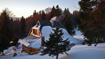 Uma cabana coberta de neve com telhado inclinado, rodeada por árvores e uma paisagem nevada.