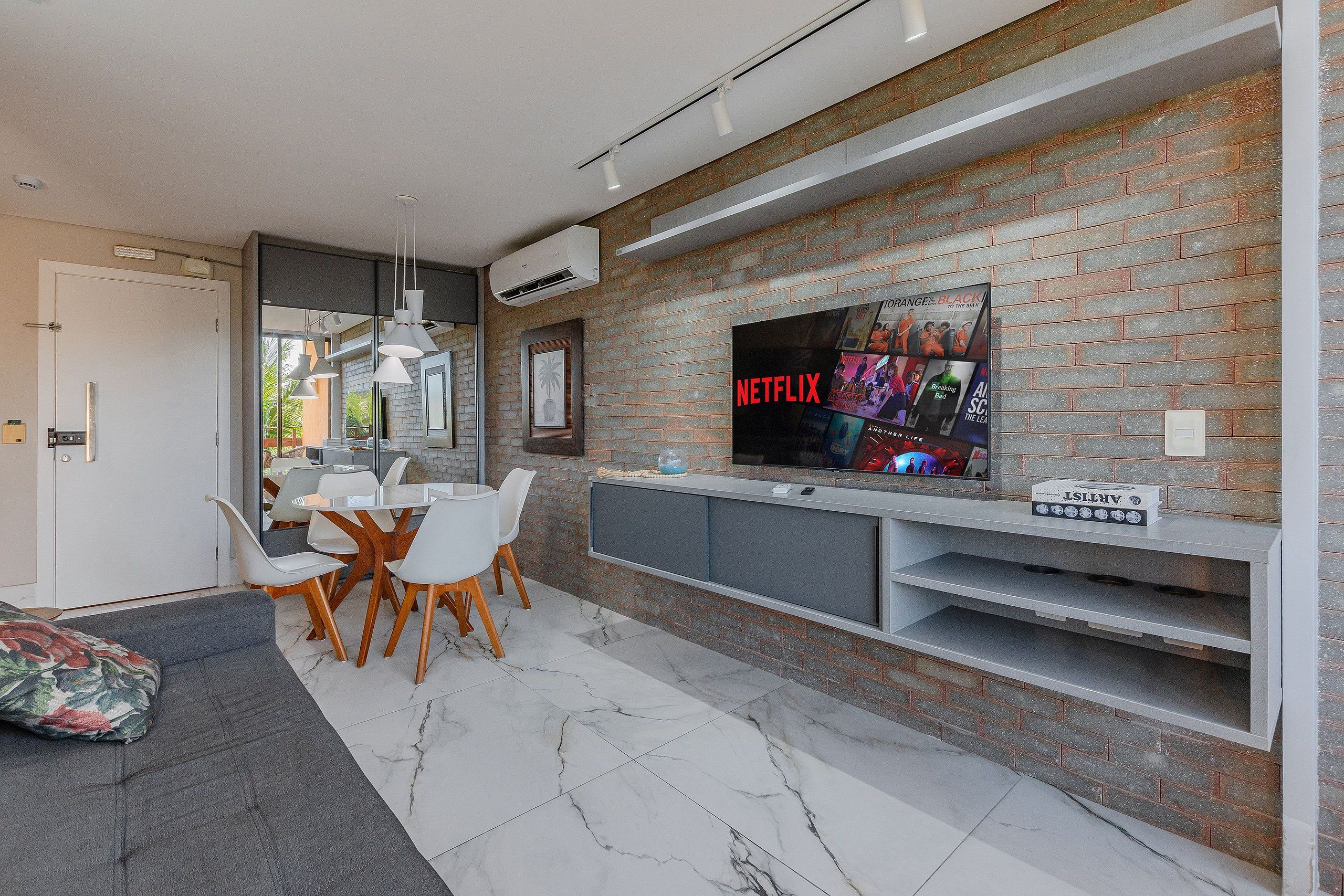 Uma sala de estar moderna com parede de tijolos, uma TV de tela plana montada acima de um console e um espaço de jantar com cadeiras brancas e mesa de vidro.