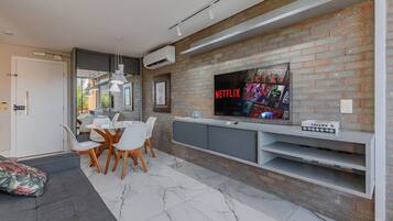 Uma sala de estar moderna com parede de tijolos, uma TV de tela plana montada acima de um console e um espaço de jantar com cadeiras brancas e mesa de vidro.