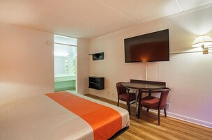 Quarto de hotel com cama, escrivaninha, cadeira, televisão e abajur.
