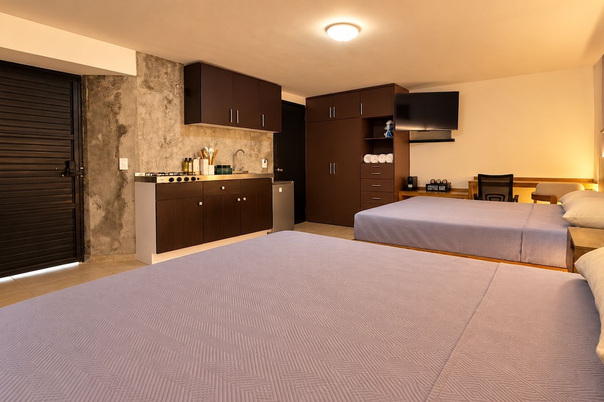 Quarto de hotel com duas camas, uma cozinha compacta, televisão de tela plana e luminária fixada na parede.