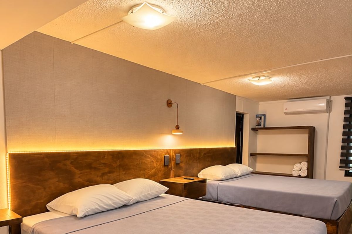 Quarto de hotel com duas camas, cabeceiras de madeira, uma mesa de cabeceira, prateleira na parede e luminária no teto.