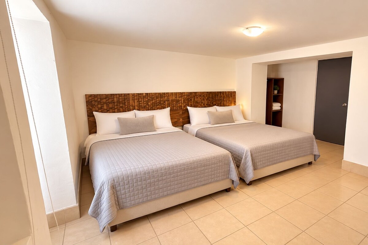 Quarto de hotel com duas camas, cabeceira de madeira e porta para um banheiro.