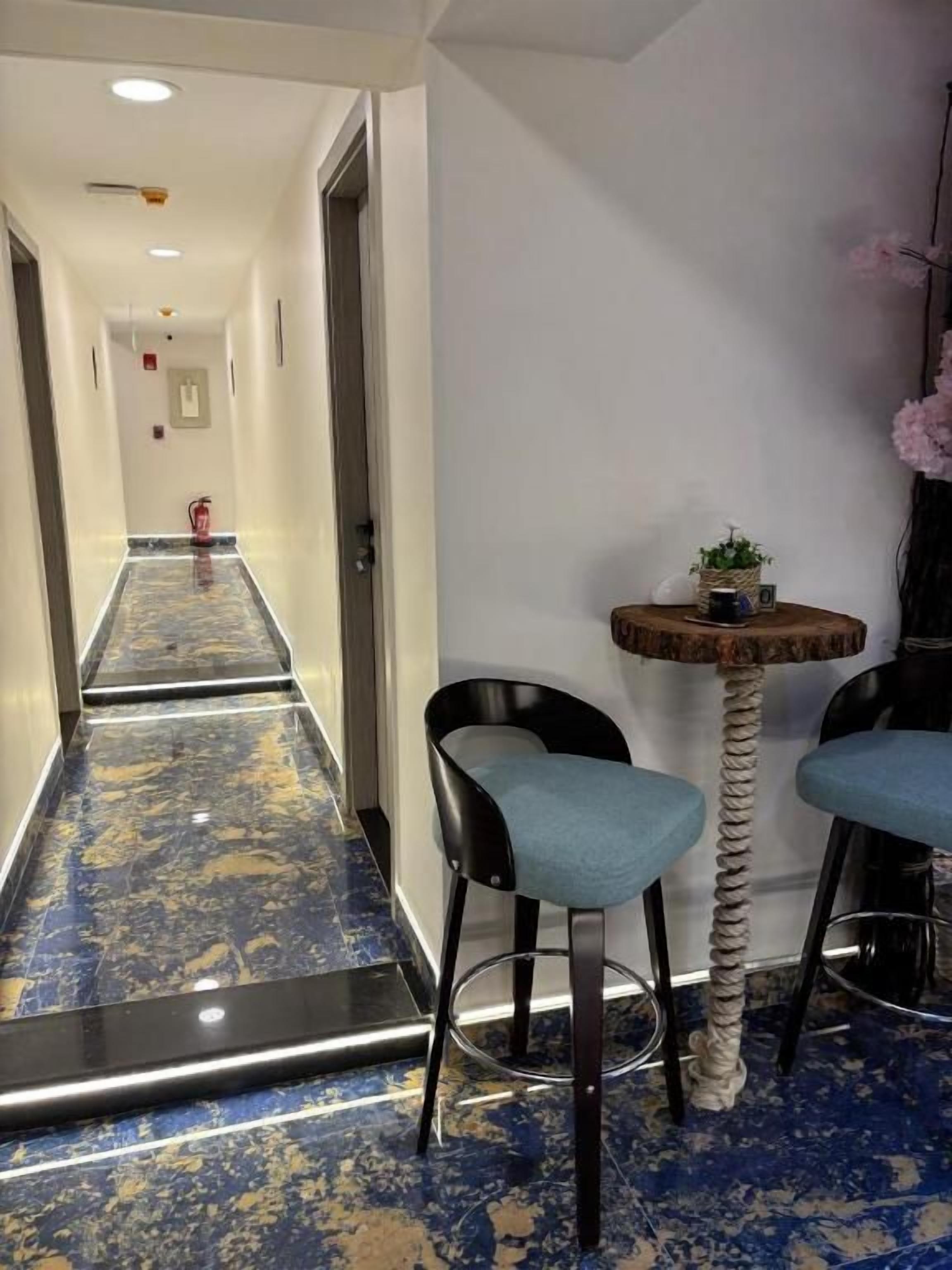 Corredor de hotel com piso estampado, uma pequena mesa redonda com uma planta e dois banquetes.