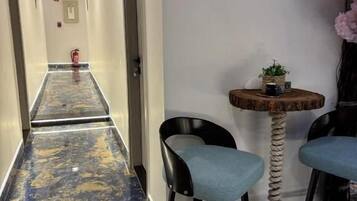 Corredor de hotel com piso estampado, uma pequena mesa redonda com uma planta e dois banquetes.