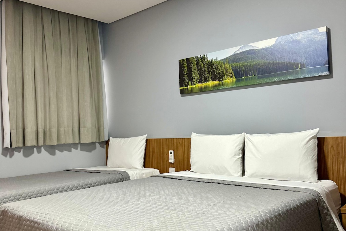 Quarto de hotel com duas camas, uma janela grande com cortinas, um quadro pendurado na parede e uma cabeceira de madeira.