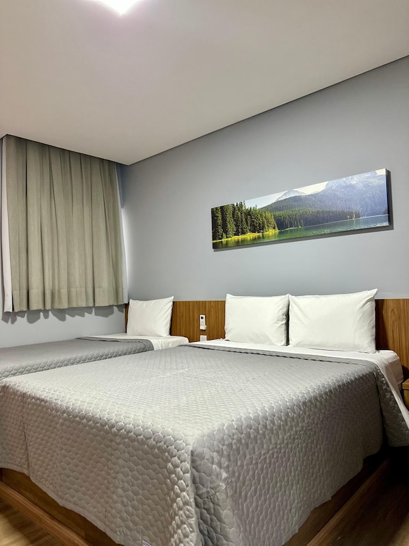 Quarto de hotel com duas camas, uma janela grande com cortinas, um quadro pendurado na parede e uma cabeceira de madeira.