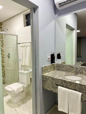 Casa de banho com chuveiro em cabine de vidro, sanita branca, lavatório com bancada de granito e espelho.
