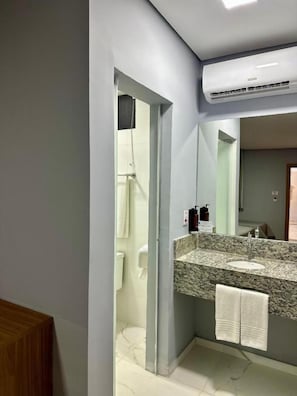 Casa de banho moderna com lavatório de bancada em granito, espelho e chuveiro visível através de uma porta de vidro.